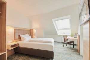 Quarto de hotel com cama, mesa de cabeceira, cadeira, escrivaninha e janela.