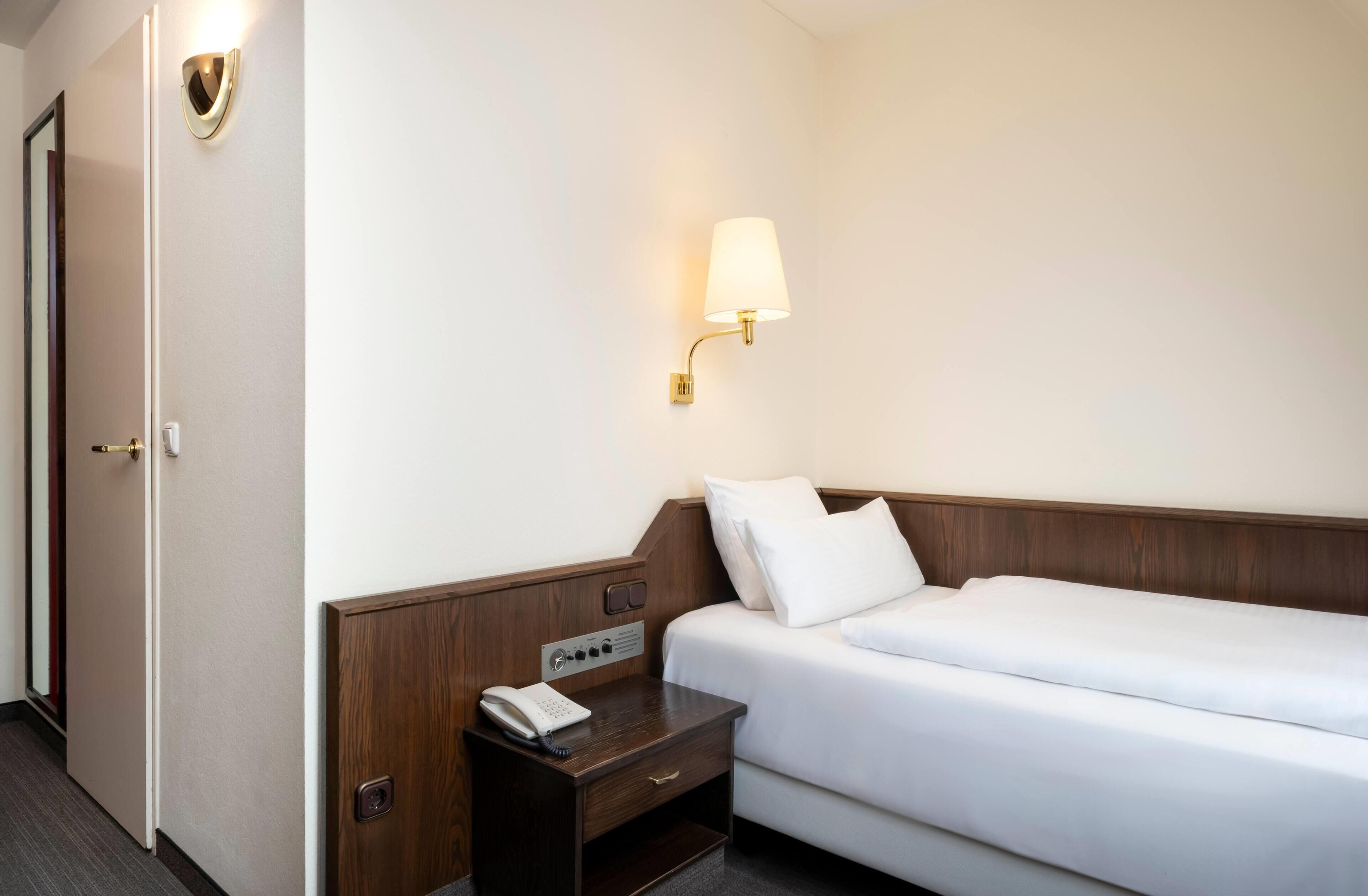 Quarto de hotel com cama, mesa de cabeceira com telefone, luminária na parede e uma porta.