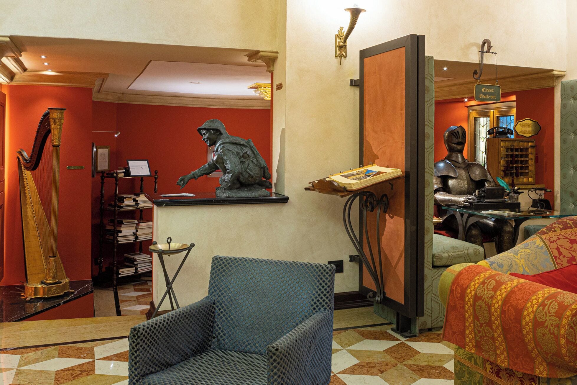 Um hall de hotel com uma harpa, uma estátua de um homem, uma mesa com um computador e uma poltrona estampada.