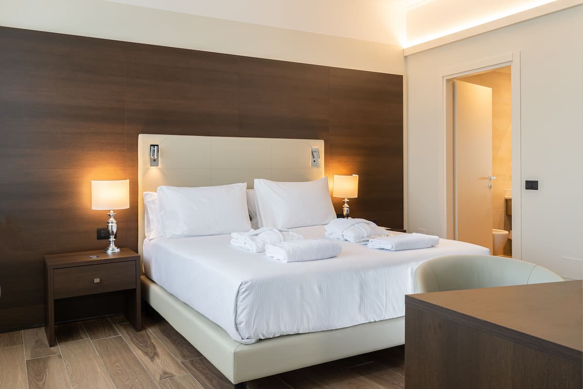 Quarto de hotel com cama, luminárias de cabeceira, uma cadeira e uma escrivaninha.