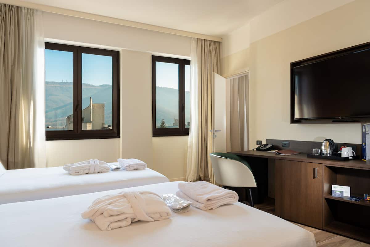 Quarto de hotel com duas camas, uma mesa, uma televisão e vista para as montanhas.