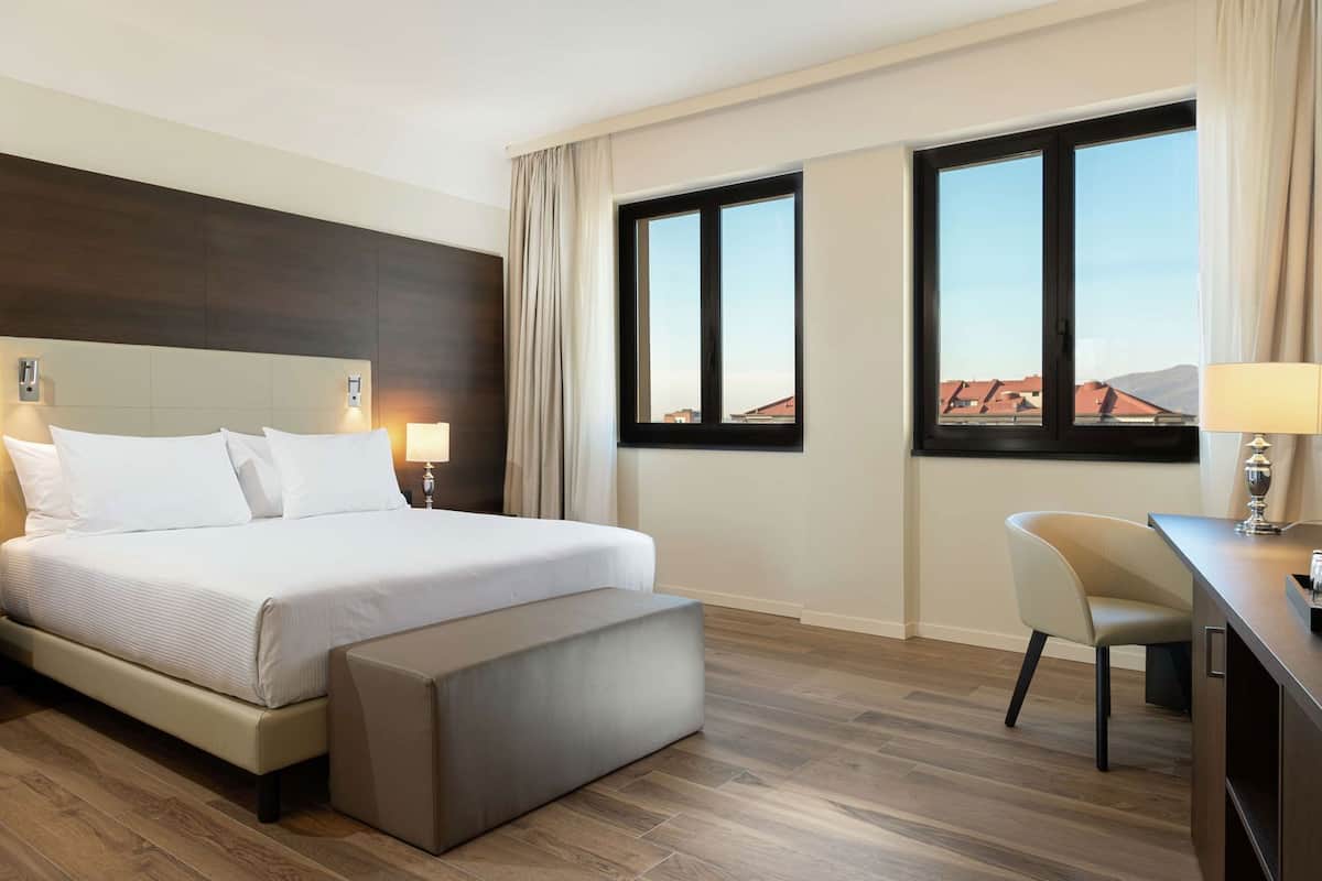 Quarto de hotel com uma cama grande, uma escrivaninha, uma cadeira e vista para edifícios pelas janelas.
