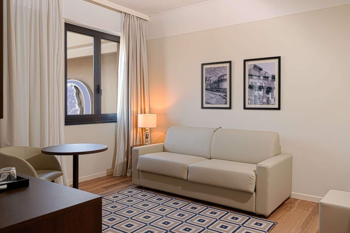 Quarto de hotel com sofá bege, uma mesinha redonda, luminária e duas fotos emolduradas na parede.