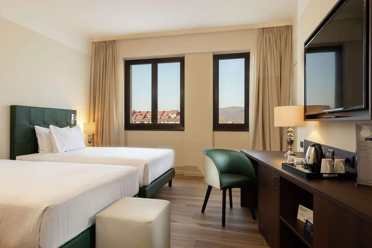 Quarto de hotel com duas camas, uma escrivaninha, uma cadeira, uma televisão e vista para a paisagem urbana.