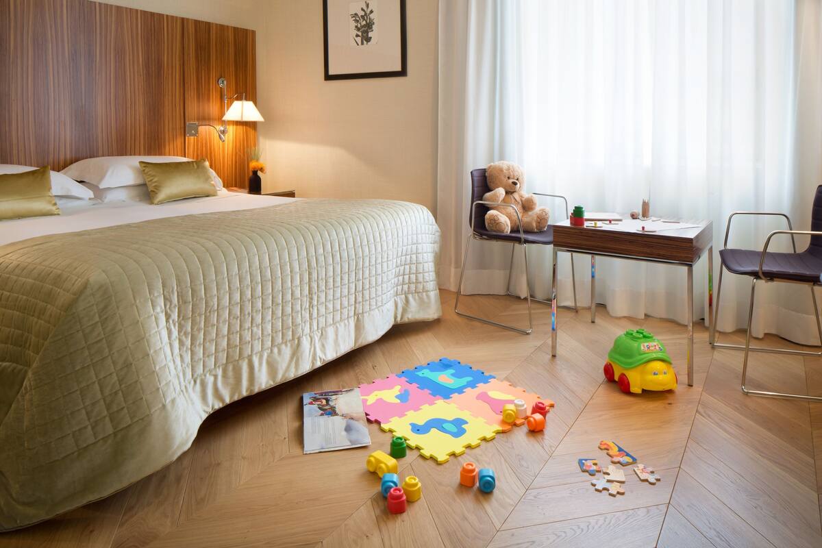Quarto de hotel com cama, escrivaninha, cadeira, um urso de pelúcia e brinquedos infantis no chão.