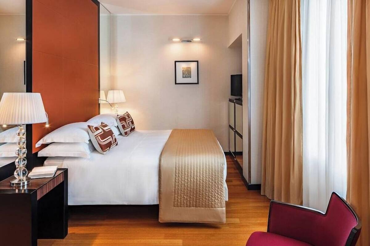 Quarto de hotel com cama, luminárias laterais, uma cadeira vermelha, televisão e janela com cortinas.