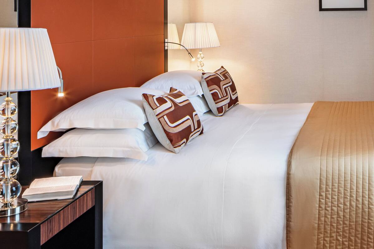 Cama bem arrumada com almofadas brancas e estampadas, mesa de cabeceira com abajur e um quadro na parede.