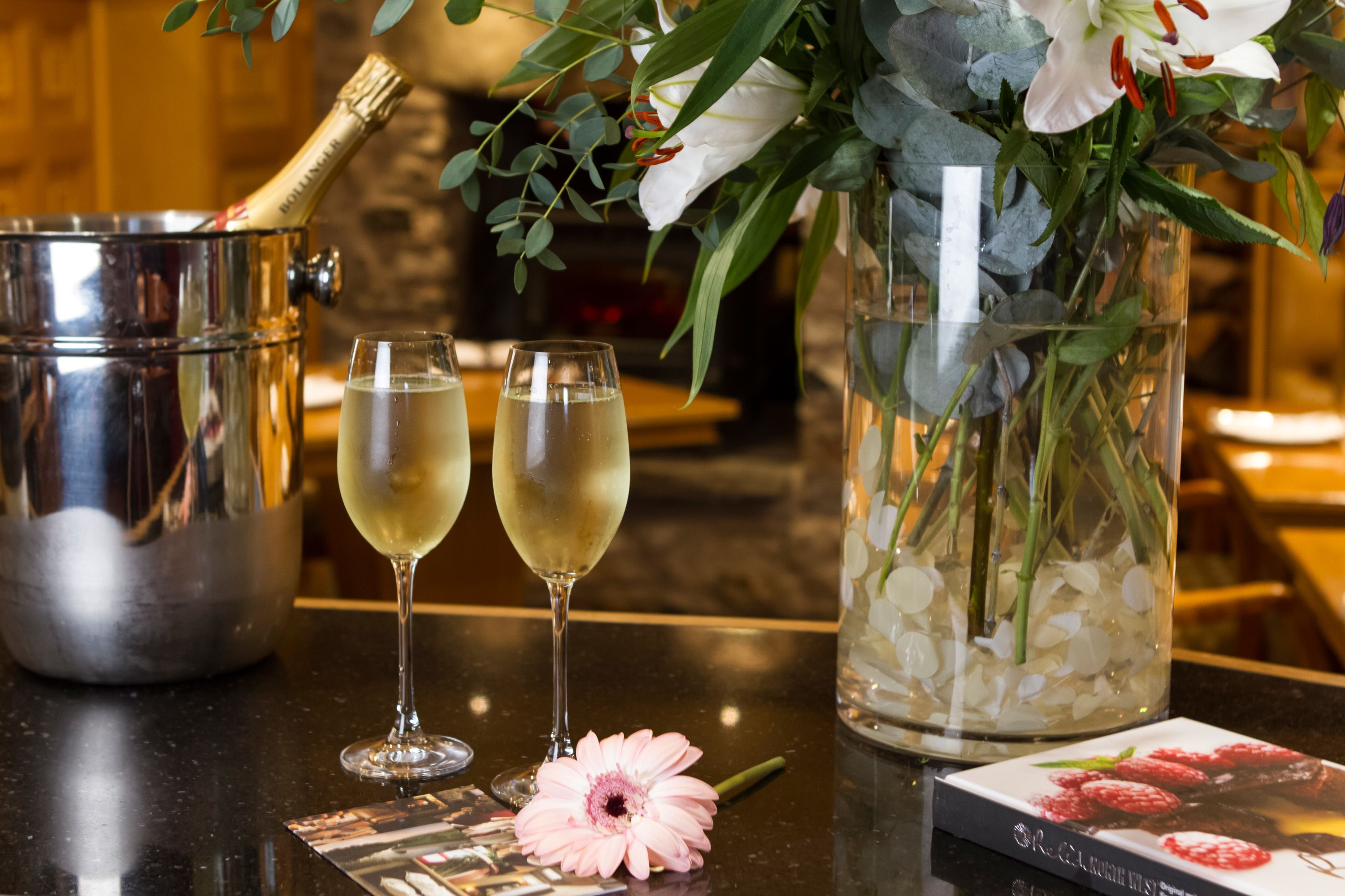 Uma área de bar com dois copos de vinho branco, uma garrafa de champanhe e um vaso com flores.