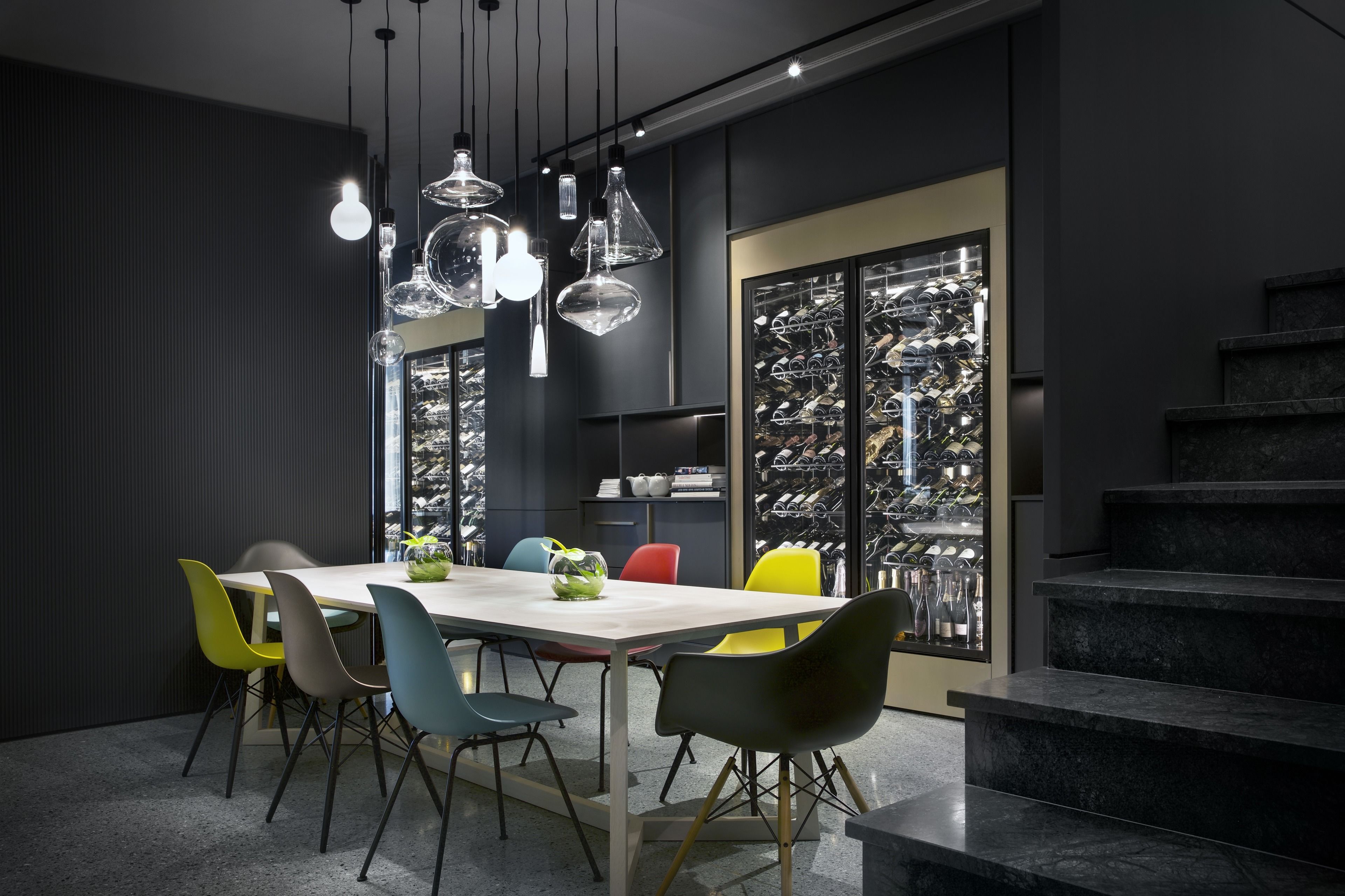 Uma sala de jantar moderna com uma mesa comprida, cadeiras coloridas e luminárias pendentes.