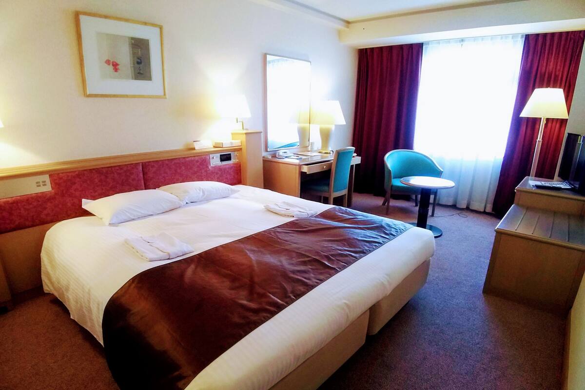 Quarto de hotel com uma cama grande, uma escrivaninha com cadeira, uma mesinha e uma televisão.