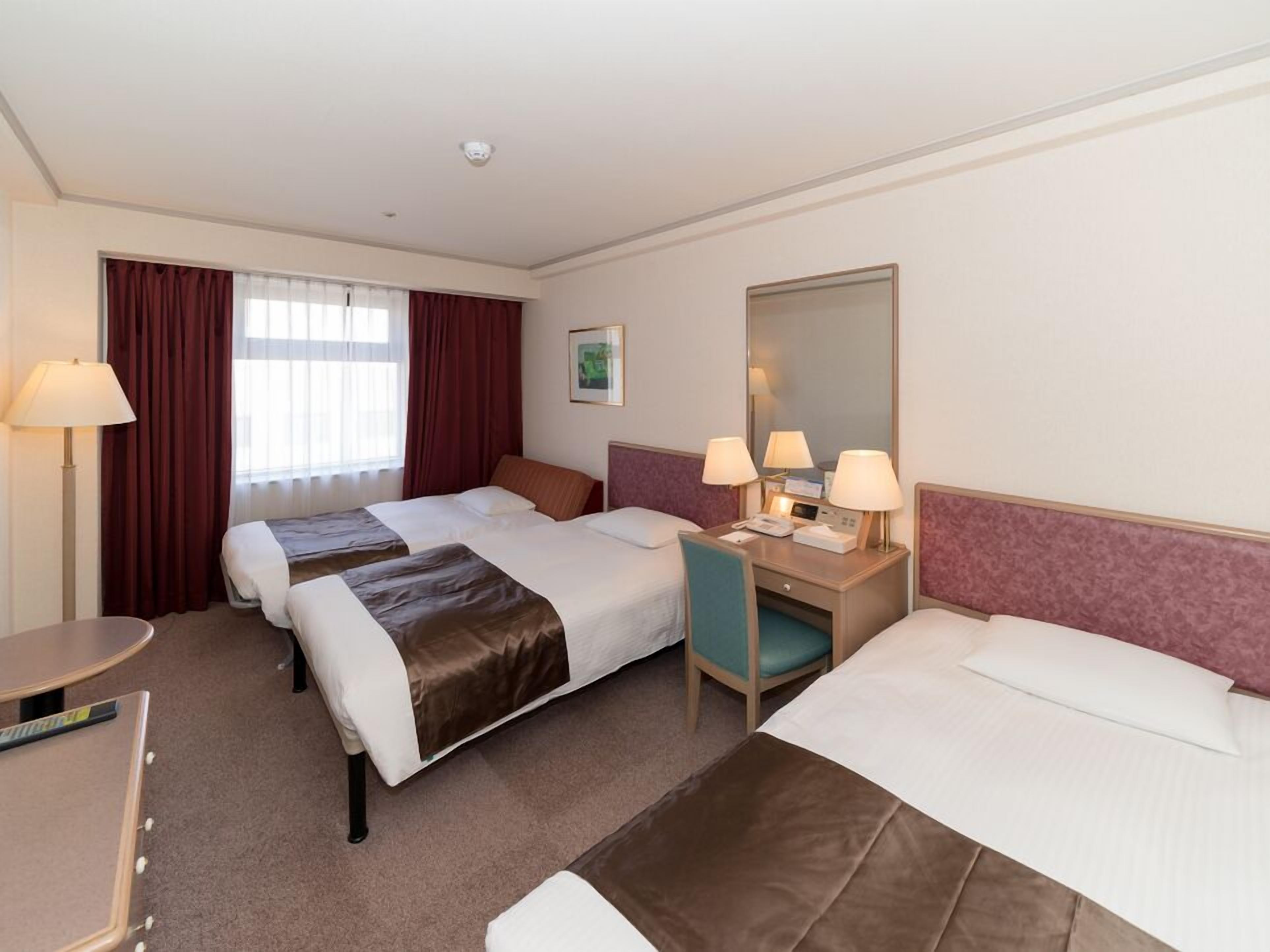 Quarto de hotel com duas camas, uma escrivaninha com cadeira, uma luminária e uma janela com cortinas.