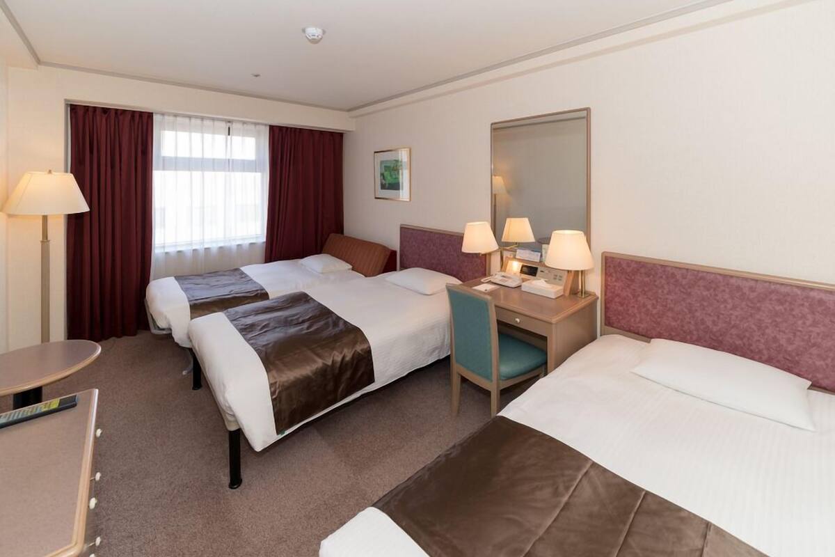 Quarto de hotel com duas camas, uma escrivaninha com cadeira, uma luminária e uma janela com cortinas.