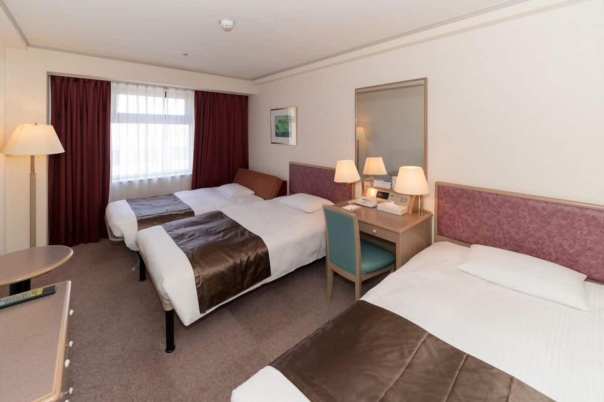 Quarto de hotel com duas camas, uma escrivaninha com cadeira, uma luminária e uma janela com cortinas.
