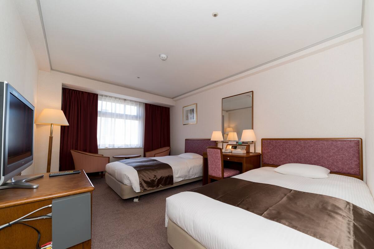 Quarto de hotel com duas camas, uma mesa com computador, televisão e janela com cortinas.
