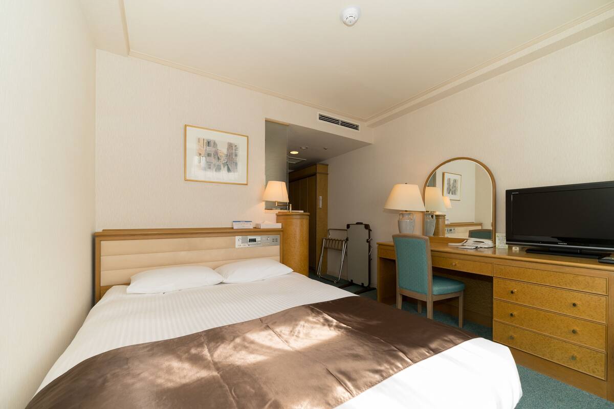 Quarto de hotel com uma cama, uma escrivaninha com cadeira, televisão e banheiro visível por uma porta aberta.