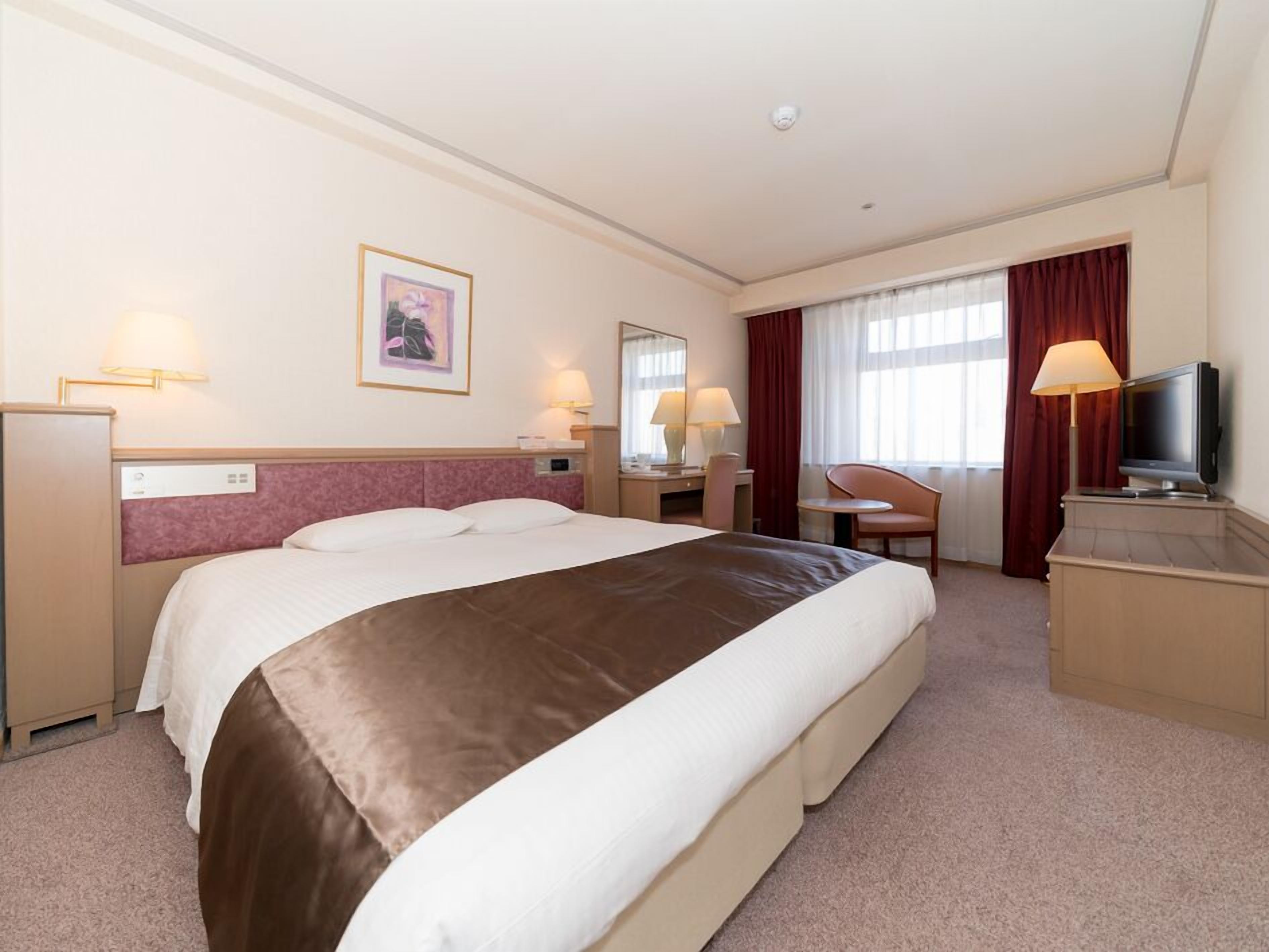 Quarto de hotel com uma cama grande, uma escrivaninha com televisão, uma cadeira e um abajur.