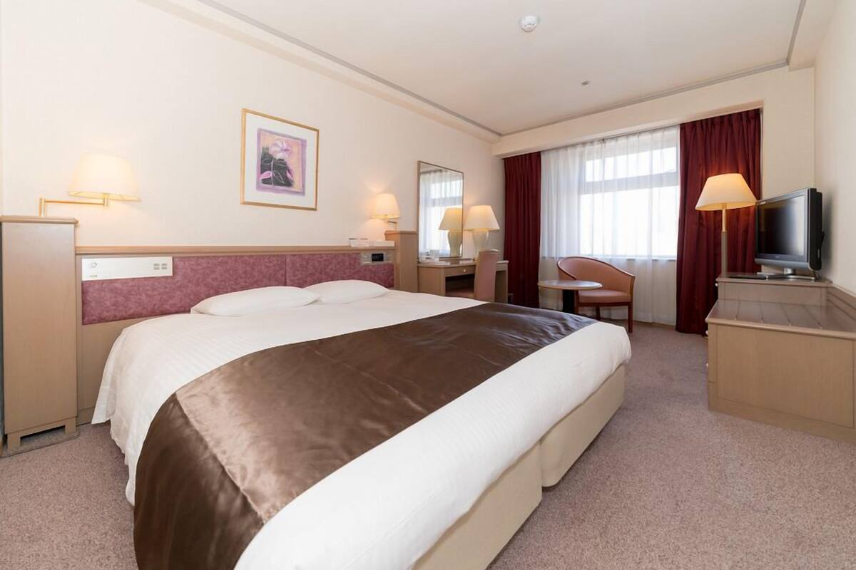 Quarto de hotel com uma cama grande, uma escrivaninha com televisão, uma cadeira e um abajur.