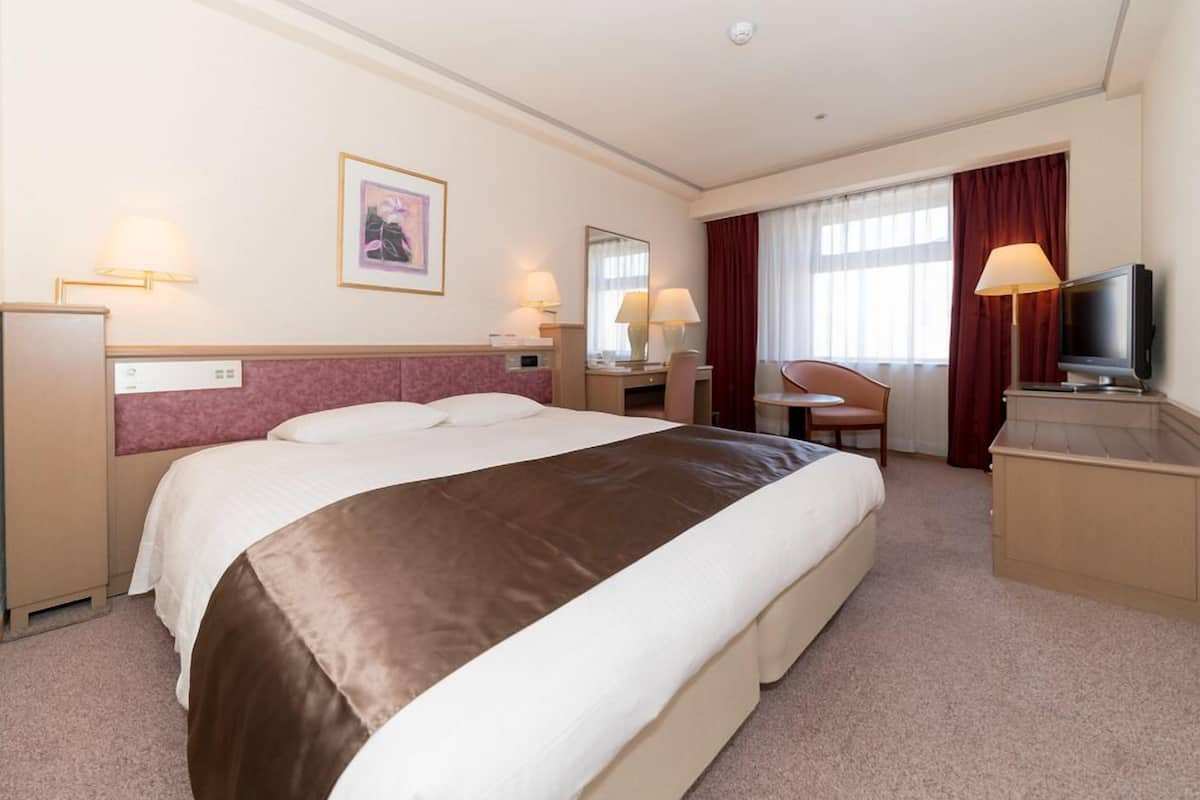 Quarto de hotel com uma cama grande, uma escrivaninha com televisão, uma cadeira e um abajur.