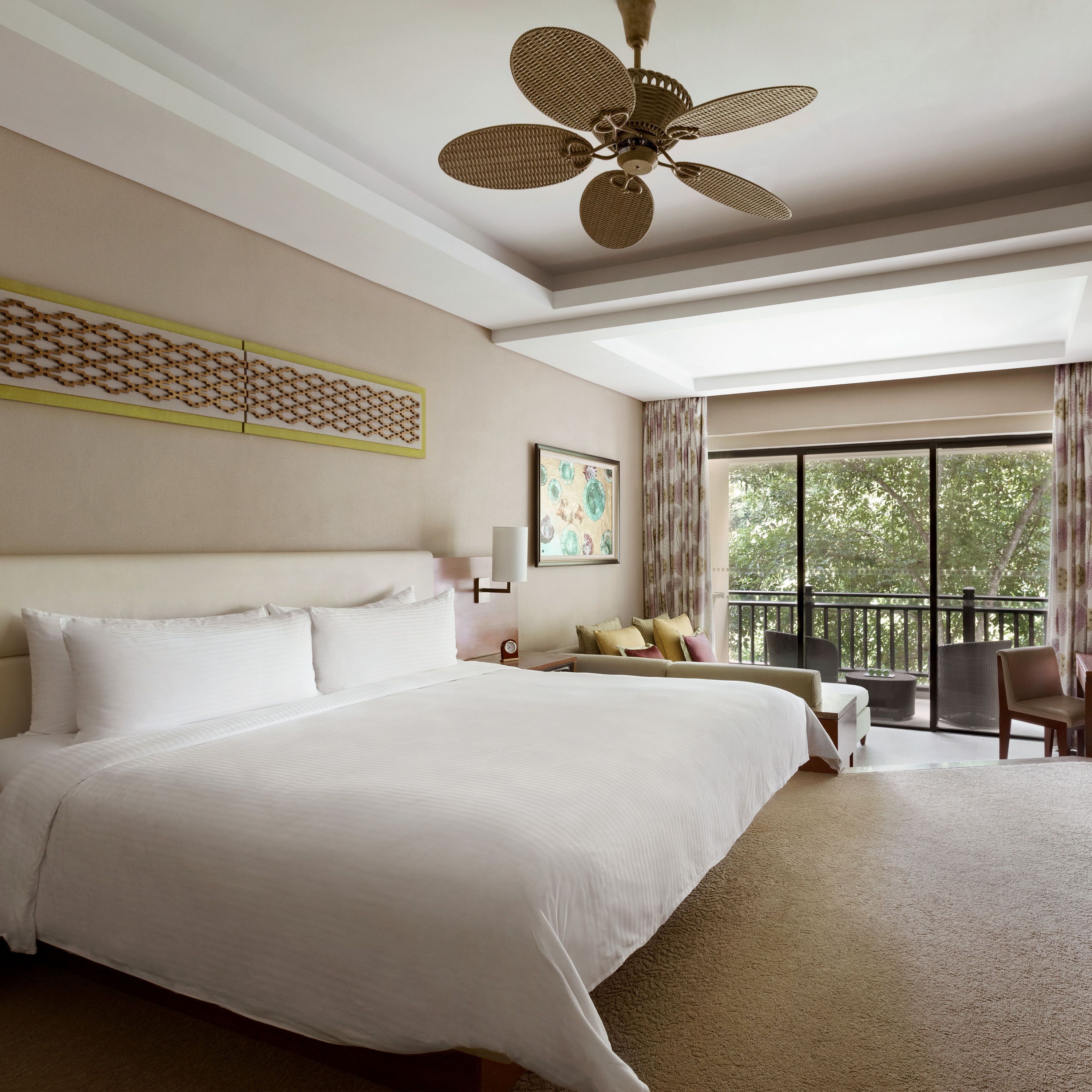 Uma cama grande com lençóis brancos, um ventilador de teto, um detalhe decorativo na parede, um quadro e uma varanda com mobiliário de exterior.