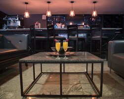 Uma área de bar com uma mesa de vidro, dois copos de suco de laranja e um balcão com garrafas e copos.