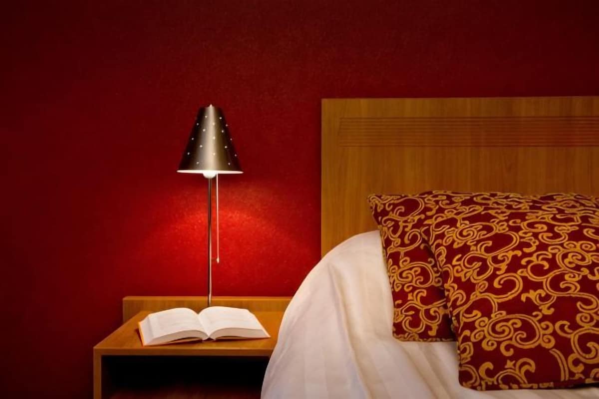 Uma cama com um travesseiro vermelho estampado, cabeceira de madeira e mesa de cabeceira com um livro aberto e uma luminária.
