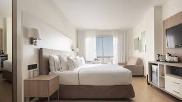 Quarto de hotel moderno com uma cama grande, mesas de cabeceira, televisão e vista para o mar.