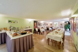 Uma sala de jantar com várias mesas preparadas para o café da manhã, um buffet com diversos alimentos e uma parede decorada com quadros.