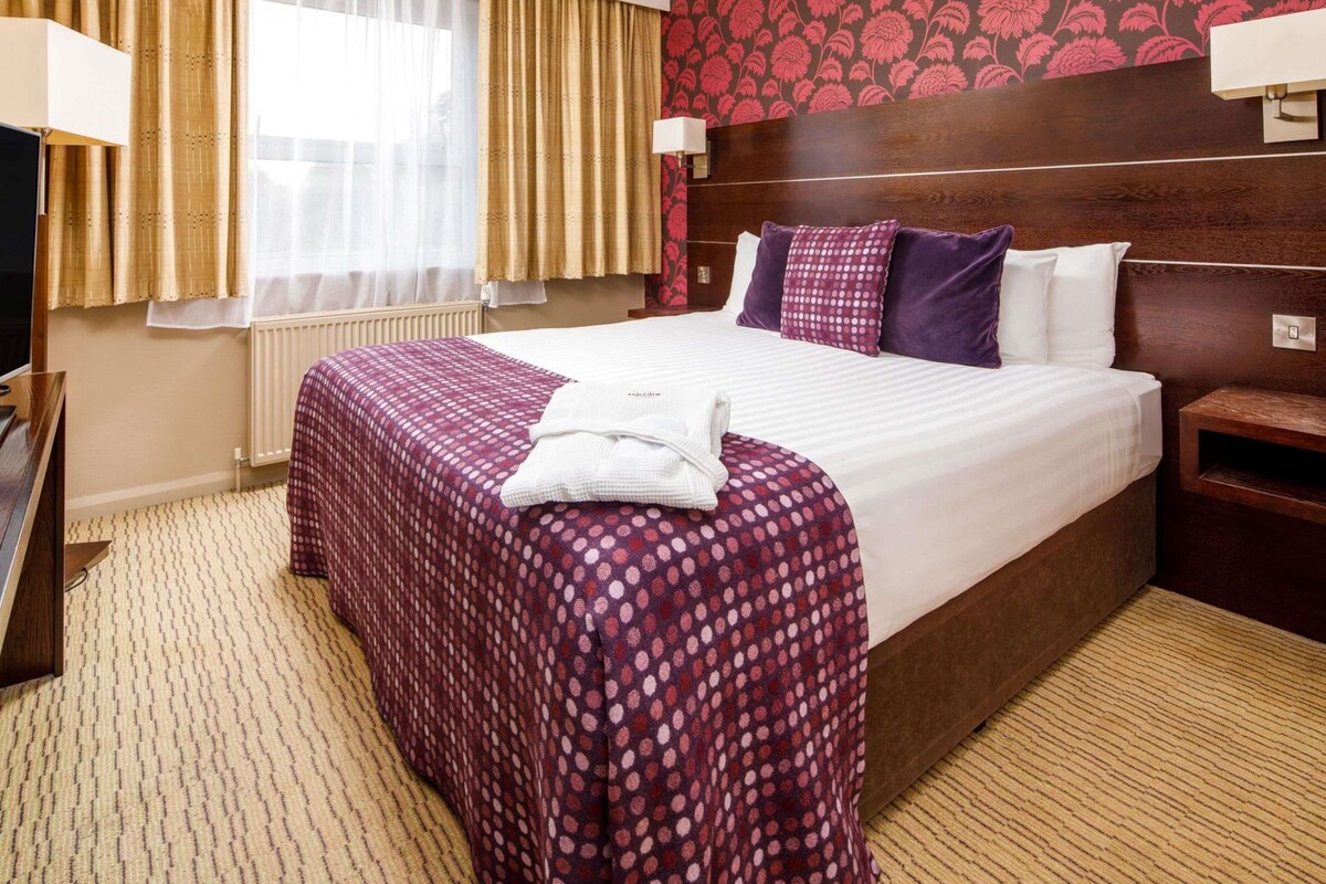 Quarto de hotel com uma cama grande, cabeceira de madeira, janela com cortinas, radiador e mesa de cabeceira com abajur.