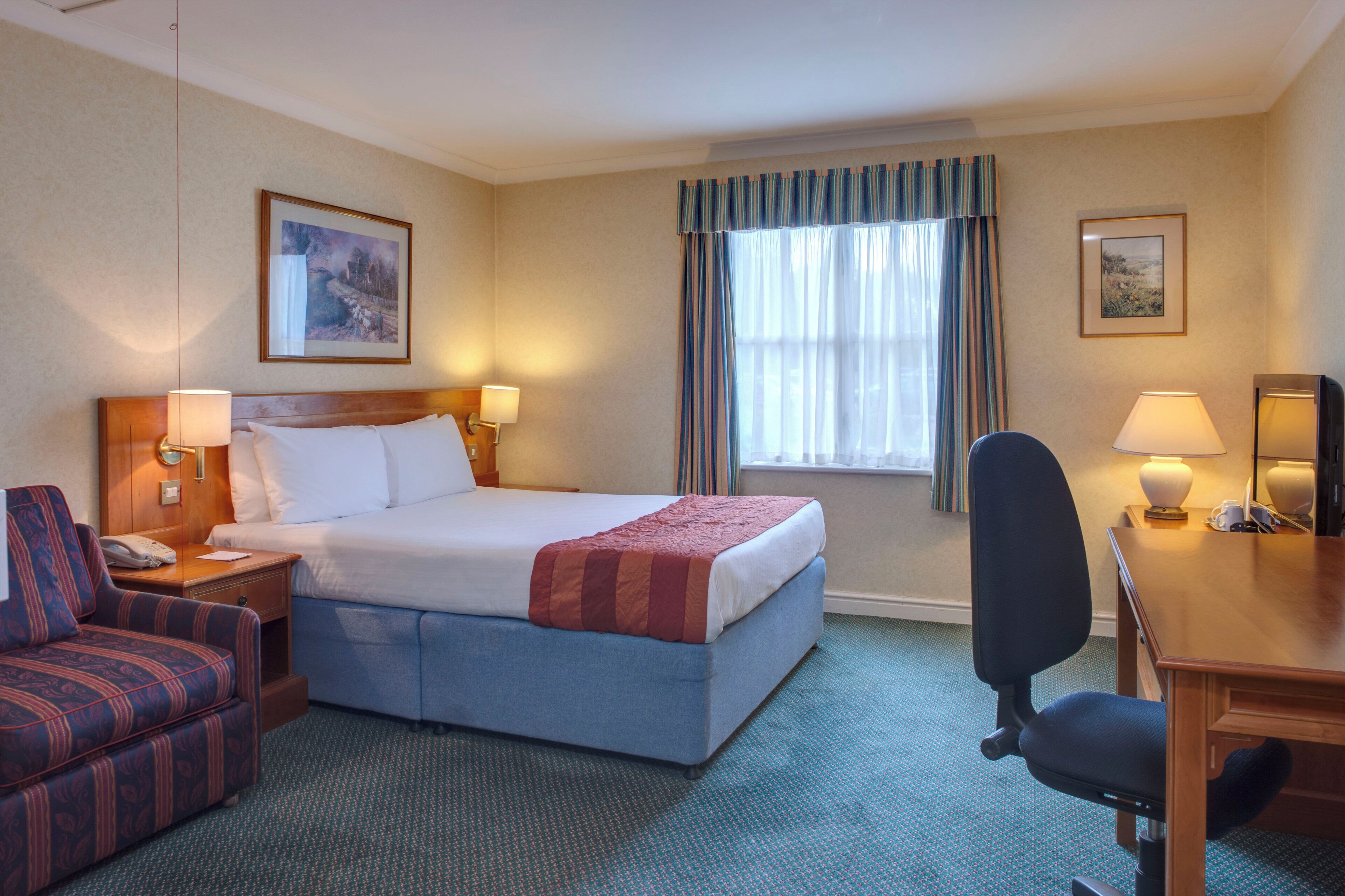 Quarto de hotel com cama, escrivaninha, cadeira, telefone e janela com cortinas.