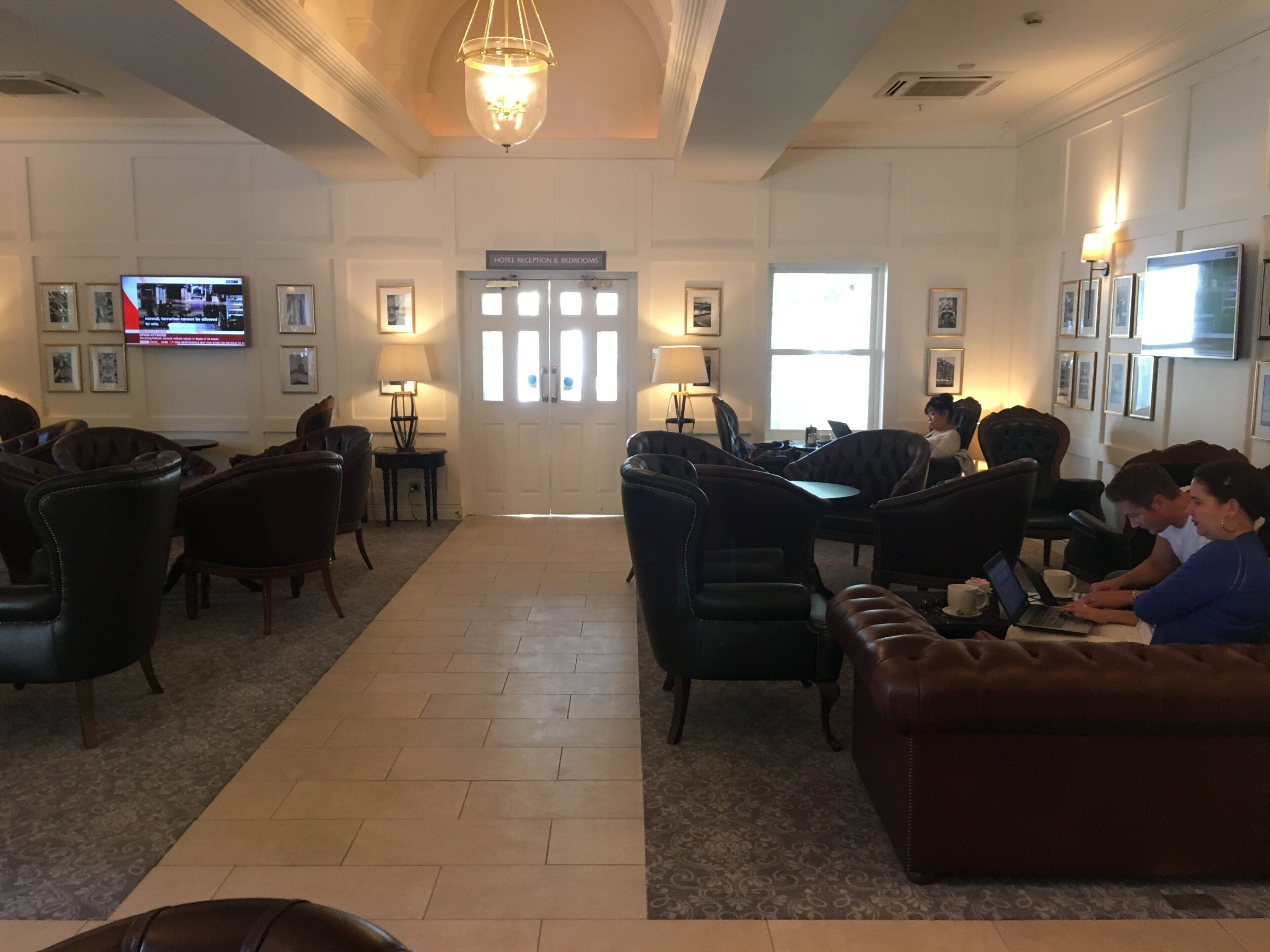 Um saguão de hotel com poltronas de couro, balcão de recepção e uma tela de televisão.