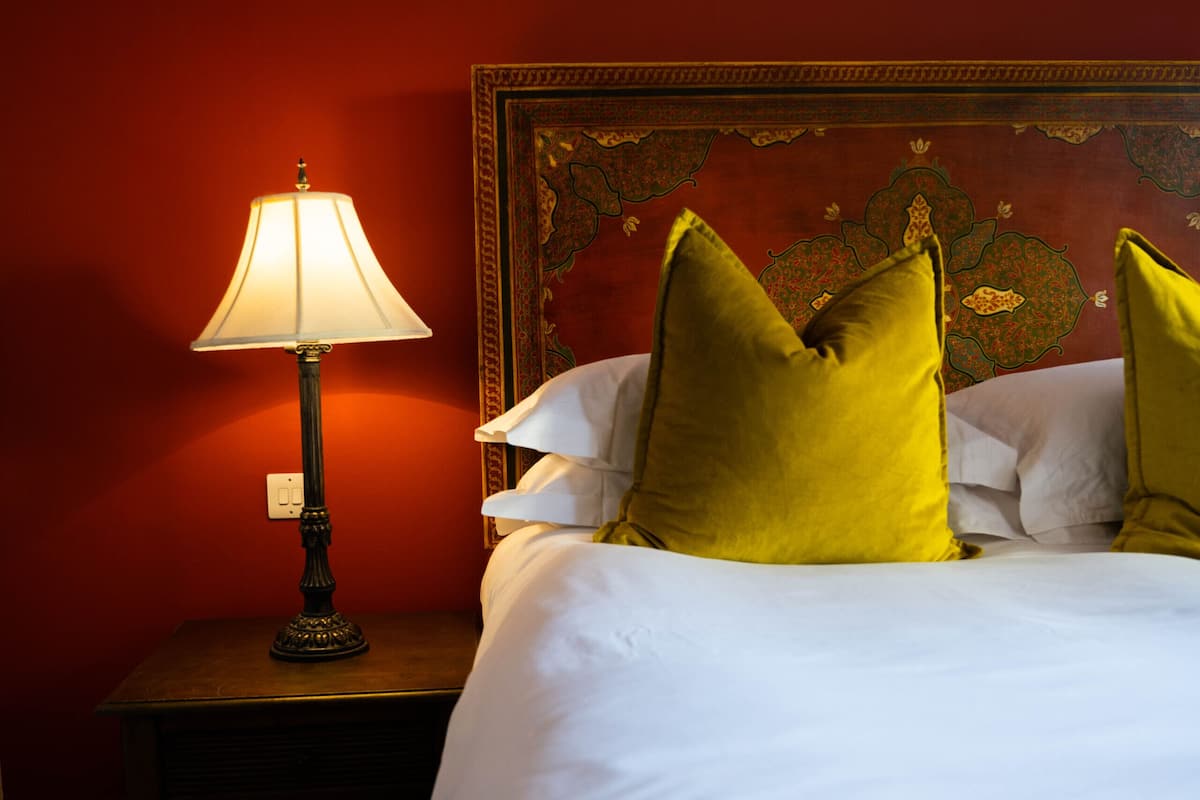 Cama bem arrumada com lençóis brancos e duas almofadas amarelas, luminária de cabeceira em mesa de madeira e parede vermelha com padrão decorativo.