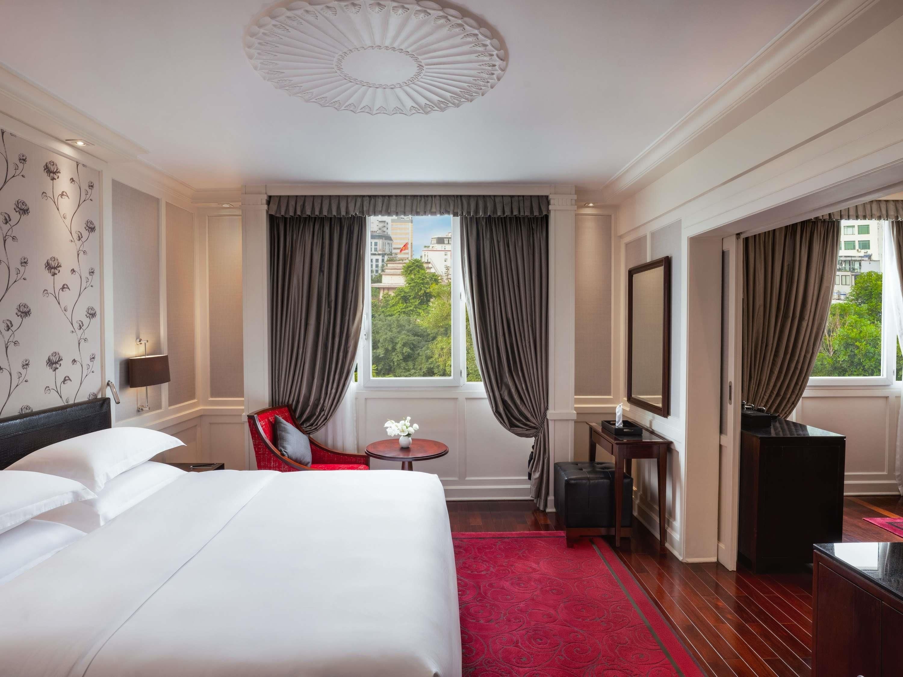 Quarto de hotel com uma cama grande, uma cadeira vermelha, uma mesinha com um vaso, um espelho e vista da cidade pelas janelas.