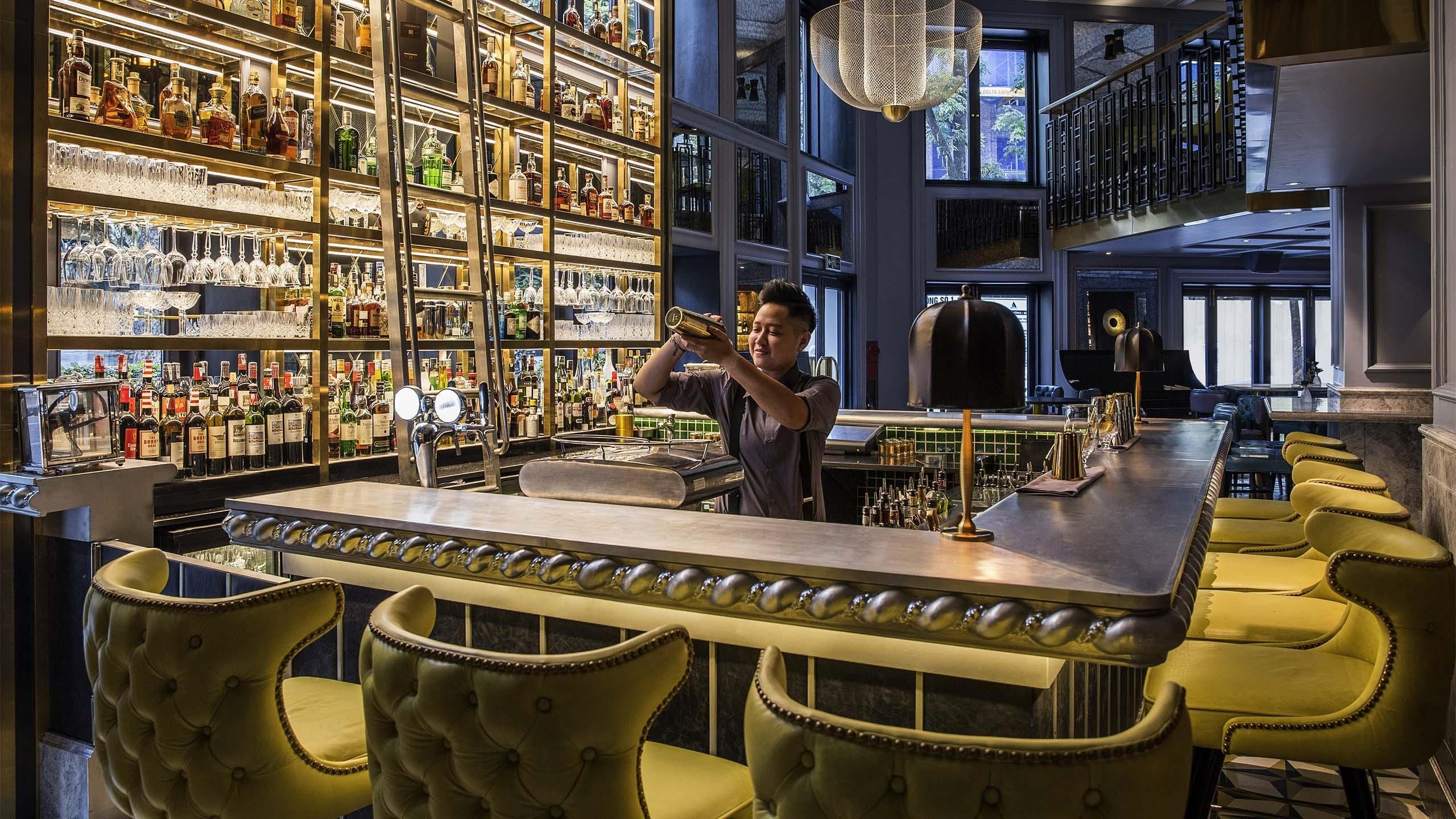 Um bar com um bartender preparando uma bebida, prateleiras de bebidas alcoólicas e cadeiras amarelas.