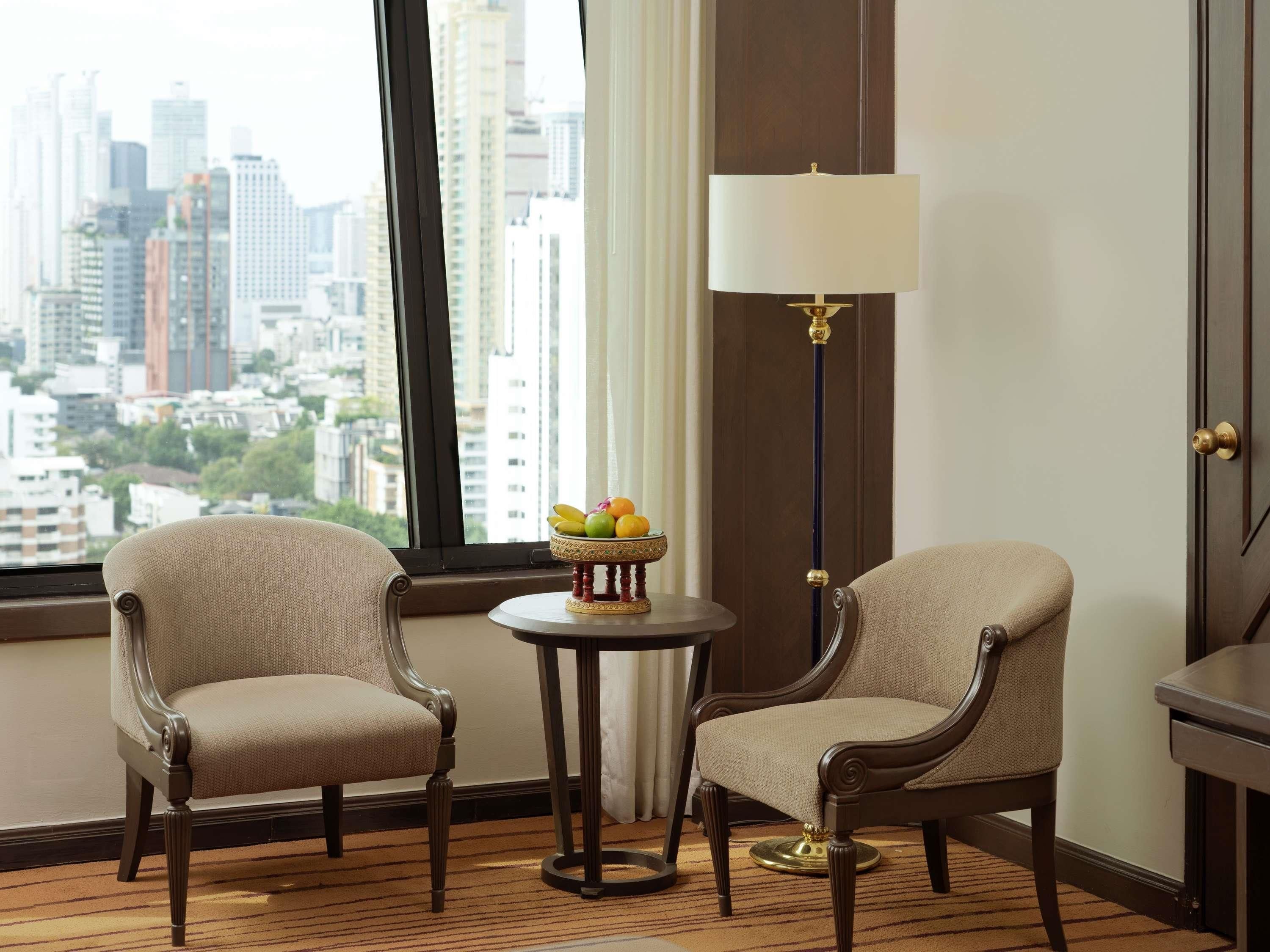 Quarto de hotel com duas poltronas, uma mesinha de centro com uma fruteira, um abajur e vista para a cidade.