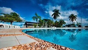 Piscina com água azul cristalina, rodeada por palmeiras e um edifício com telhado escuro.