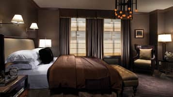 Quarto de hotel com uma cama grande, uma cadeira, uma mesa com abajur e uma janela com cortinas.