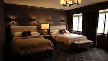 Quarto de hotel com duas camas, mesa de cabeceira, luminária e janela com cortinas.