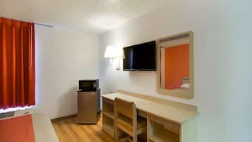 Quarto de hotel com cama, escrivaninha, cadeira, televisão, espelho, abajur e um pequeno frigorífico.