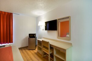 Quarto de hotel com cama, escrivaninha, cadeira, televisão, espelho, abajur e um pequeno frigorífico.