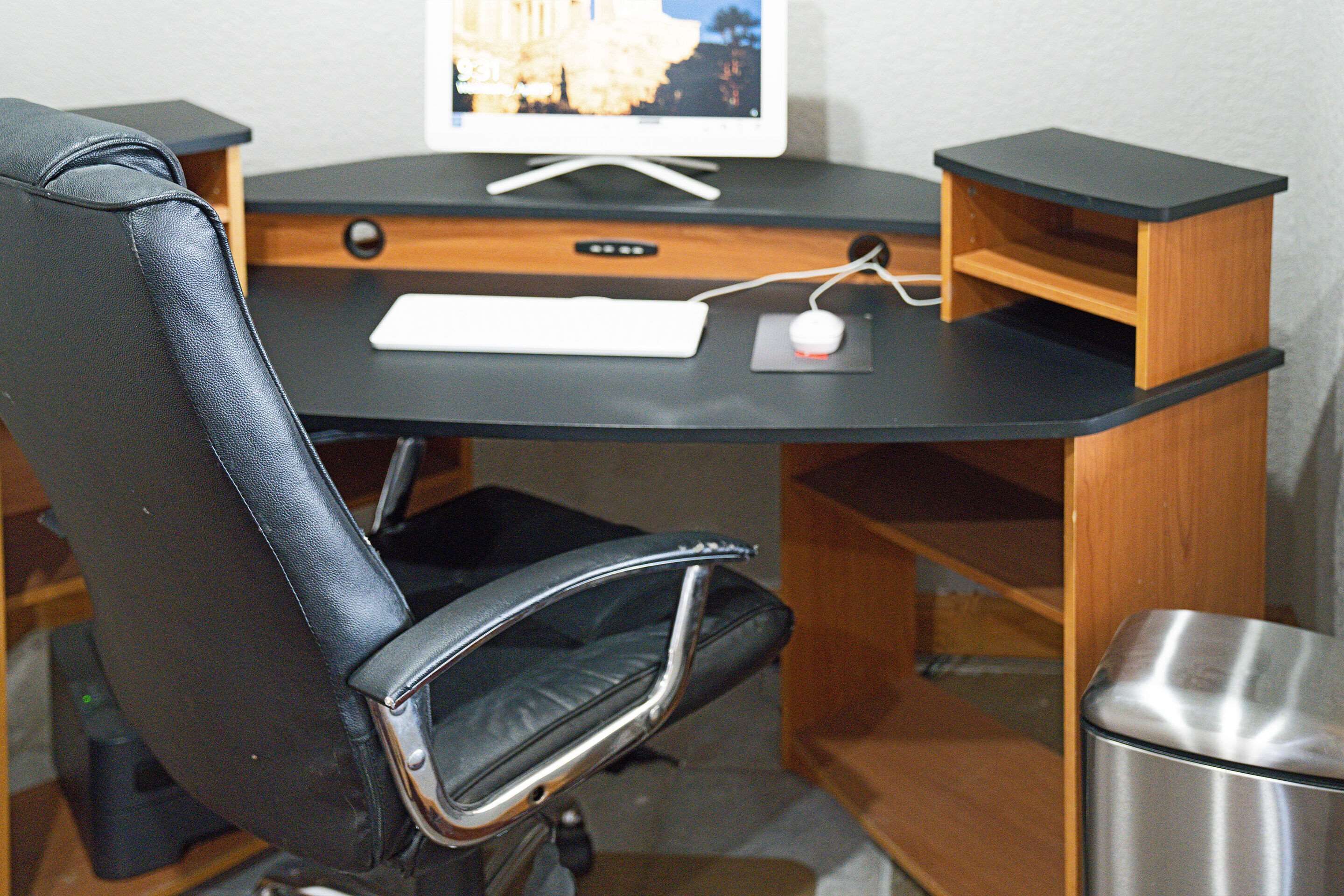 Uma cadeira de escritório preta com assento e encosto acolchoados, uma mesa de madeira com monitor, teclado e rato, e uma pequena lixeira.