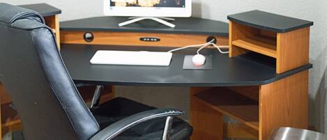 Uma cadeira de escritório preta com assento e encosto acolchoados, uma mesa de madeira com monitor, teclado e rato, e uma pequena lixeira.