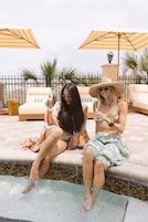 Duas pessoas sentadas em espreguiçadeiras à beira da piscina, com almofadas listradas, borda de azulejos e guarda-sóis listrados amarelos.