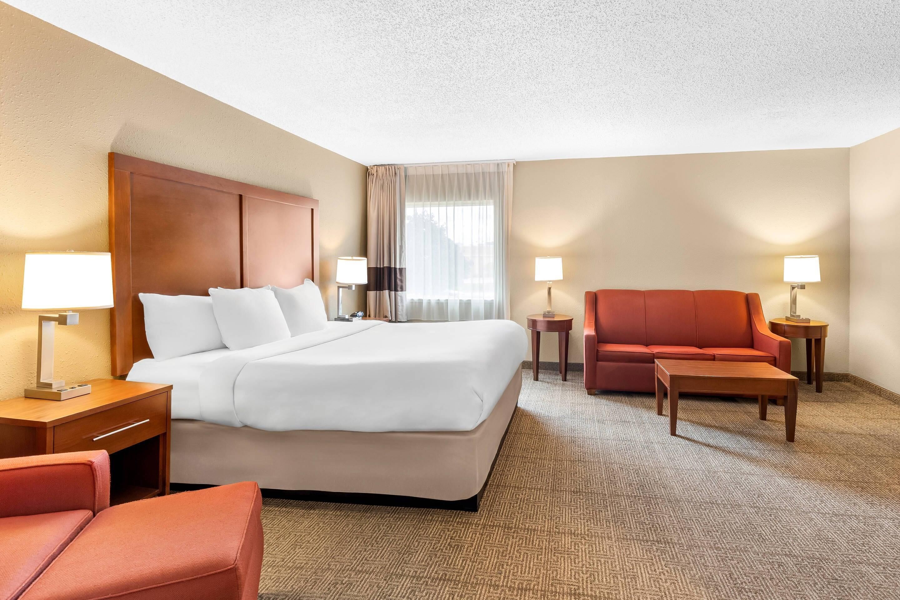 Quarto de hotel com uma cama grande, um sofá vermelho, uma mesa de centro de madeira e duas luminárias de cabeceira.