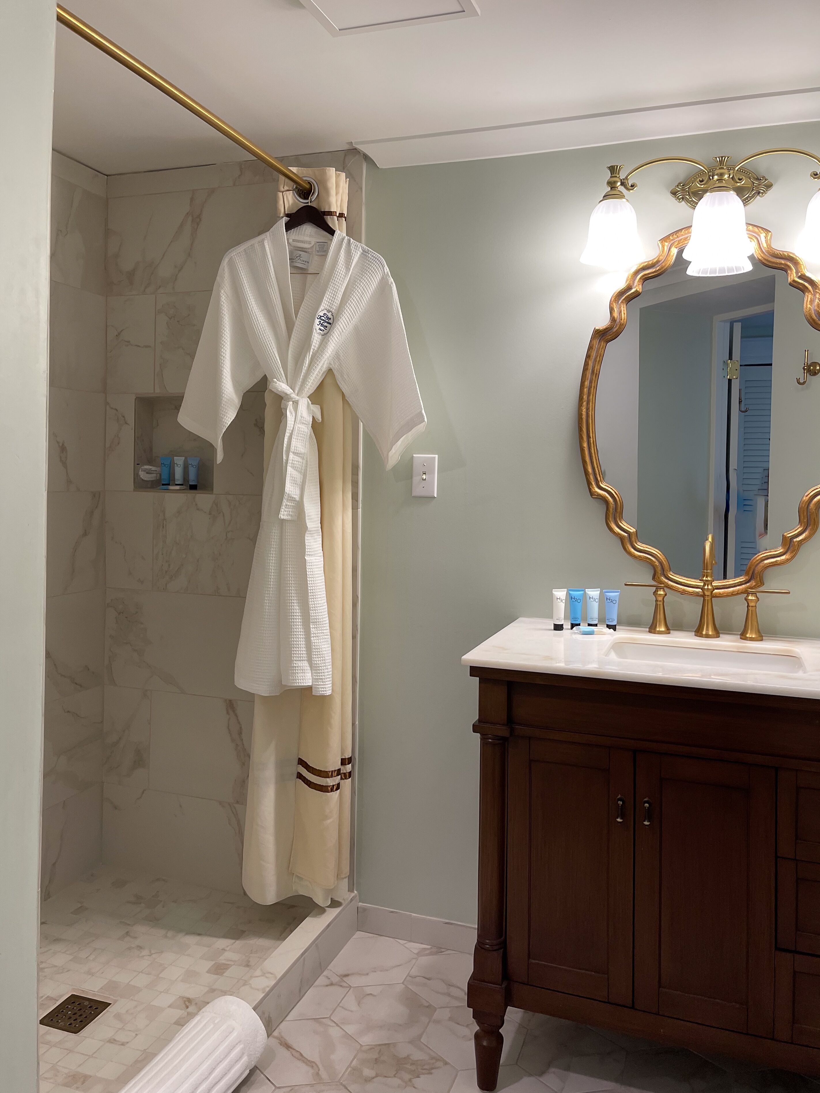 Casa de banho com chuveiro de mármore, um roupão branco pendurado num suporte, uma toucador com espelho dourado e produtos de higiene pessoal no balcão.