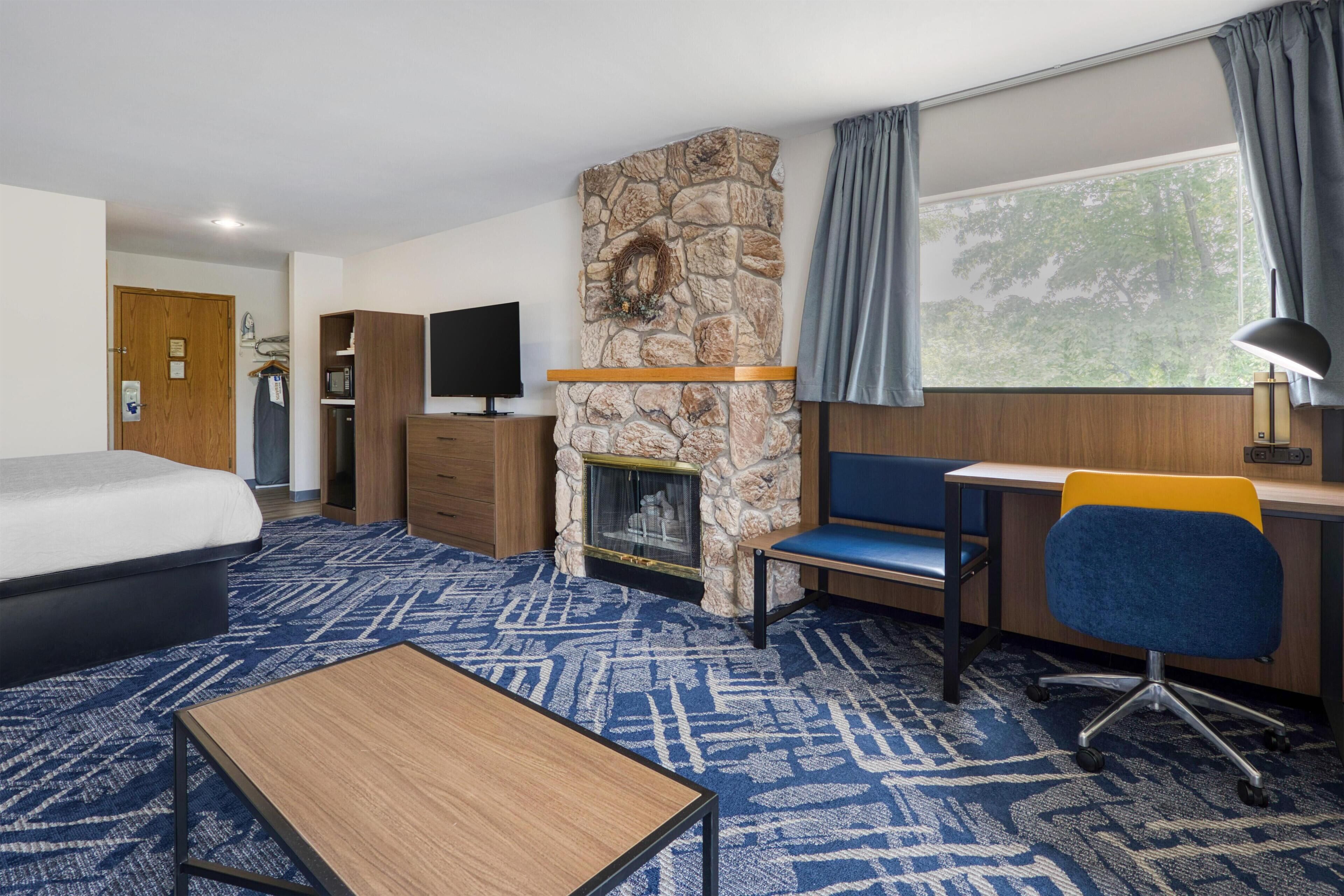 Quarto de hotel com cama, escrivaninha, cadeira, televisão, lareira de pedra e janela com cortinas.