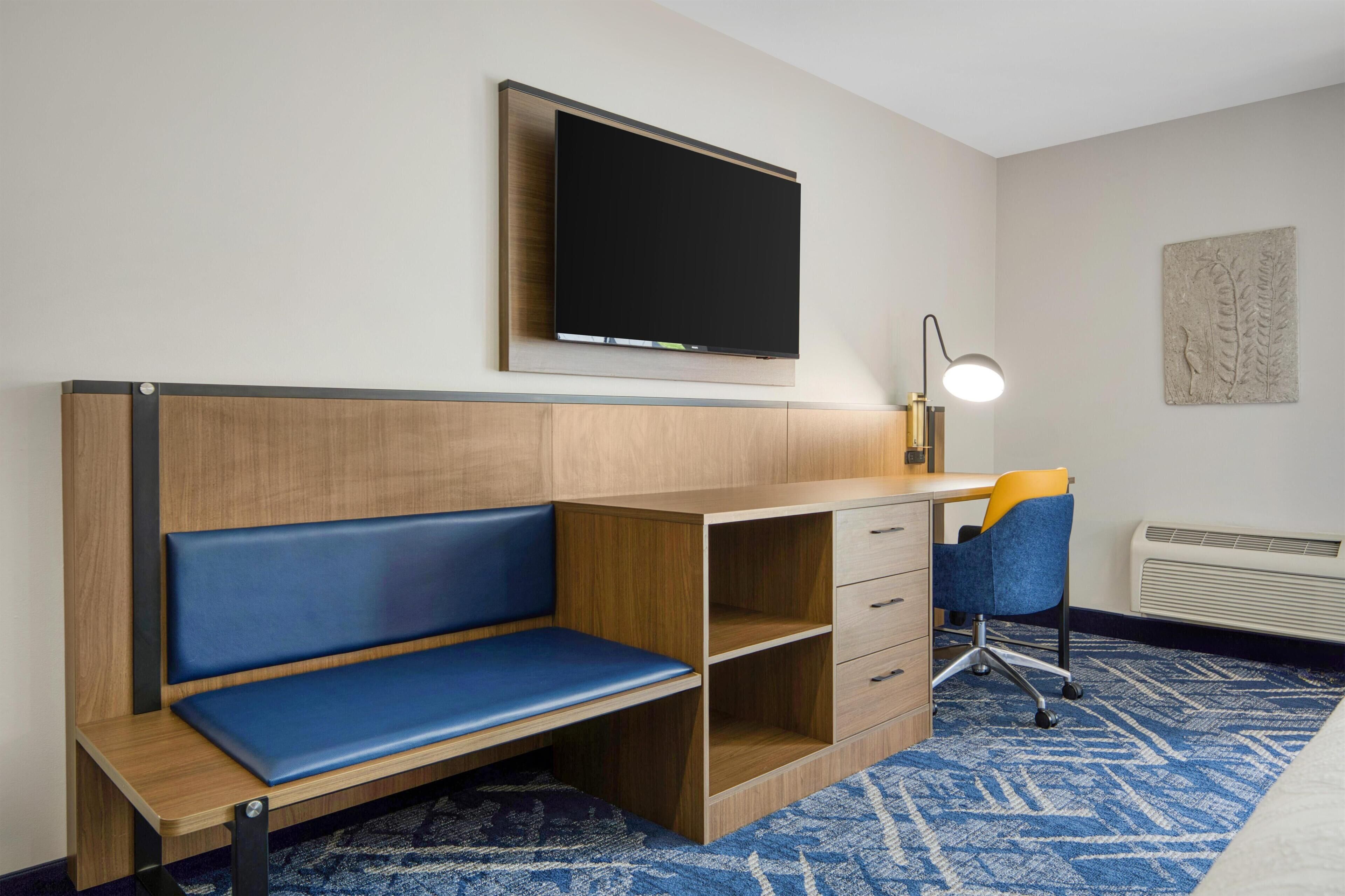 Quarto de hotel com escrivaninha de madeira, um banco azul, televisão de tela plana, luminária de mesa e radiador.