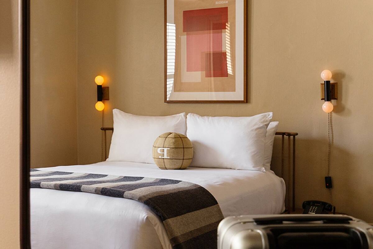 Quarto de hotel com uma cama, duas luminárias de cabeceira, uma obra de arte emoldurada e duas malas.