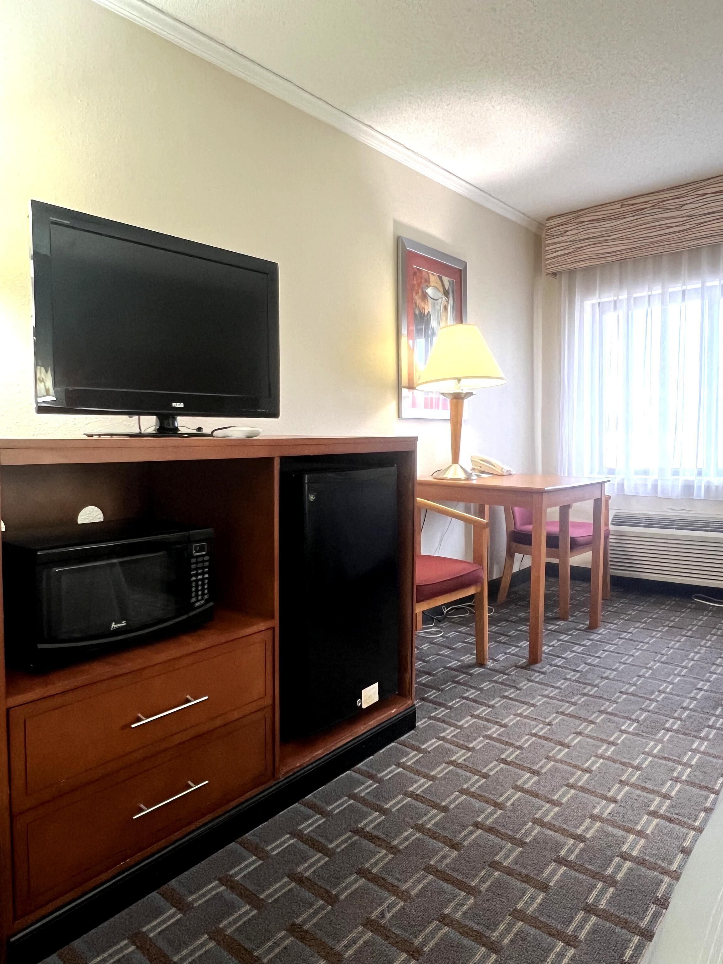 Quarto de hotel com televisão, micro-ondas e um pequeno armário. Há uma mesa de jantar com uma cadeira e um abajur.