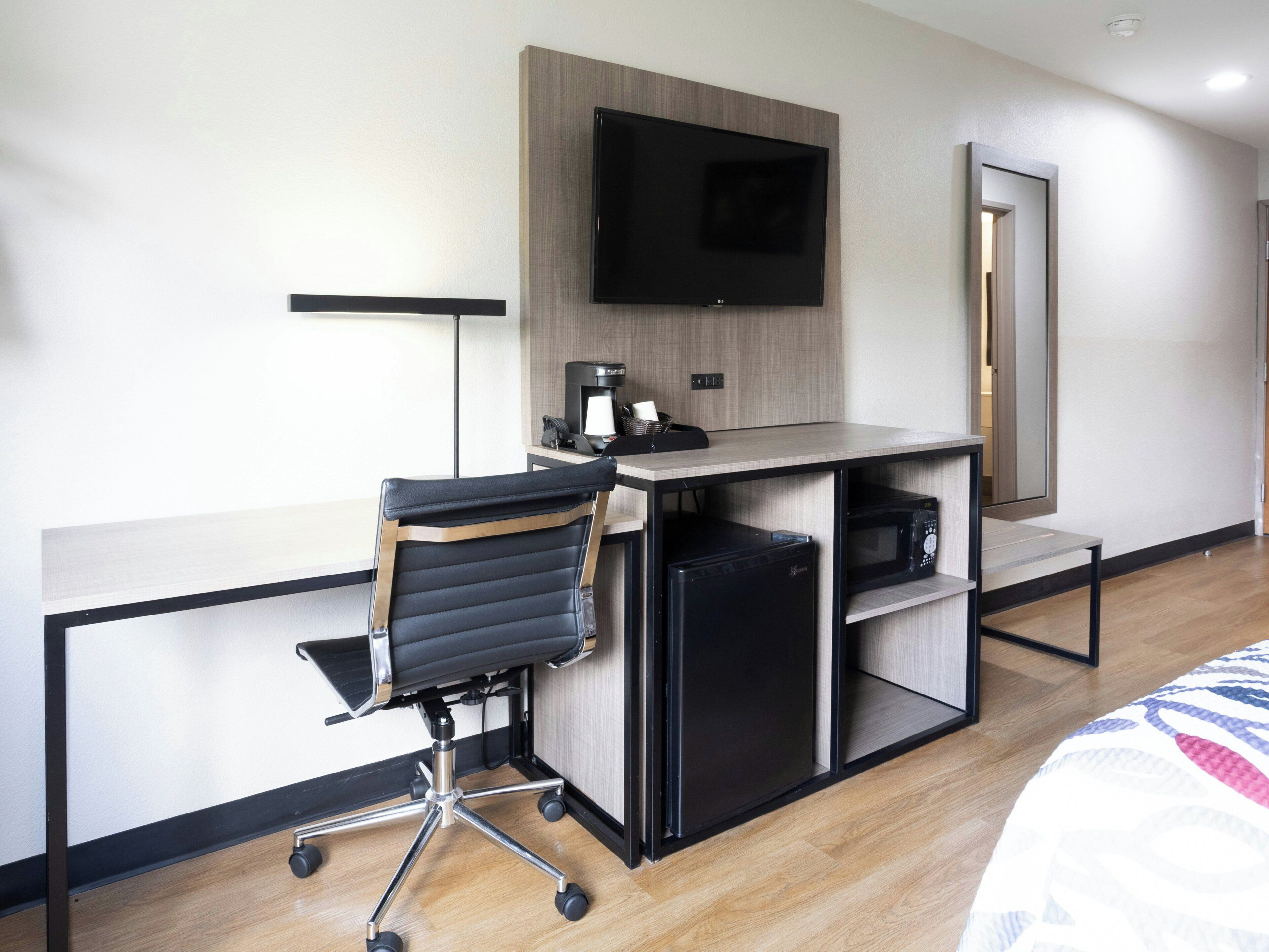 Quarto de hotel com uma escrivaninha, uma cadeira, televisão na parede, máquina de café e micro-ondas.