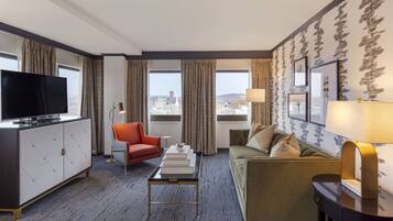 Quarto de hotel com televisão de tela grande, sofá, poltrona vermelha, mesa de centro e vista da cidade.